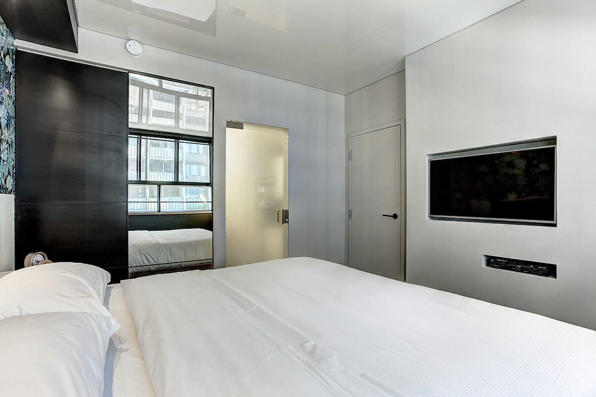Quarto de hotel moderno com uma cama grande, um guarda-roupa com espelho, uma televisão na parede e uma janela que permite a entrada de luz natural.