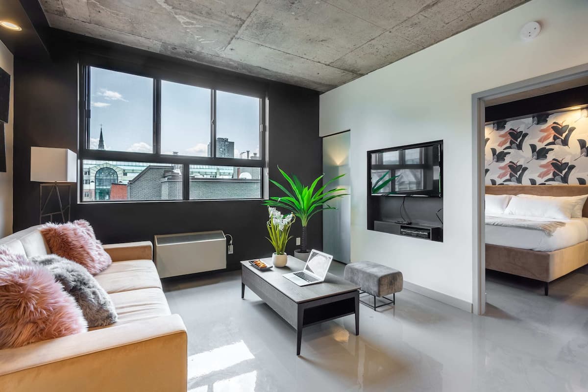 Uma sala de estar moderna com sofá, mesa de centro e um laptop. Há uma janela grande com vista para a cidade, uma planta em vaso e uma cama com cabeceira estampada.