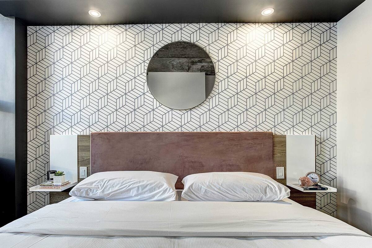 Quarto moderno com papel de parede estampado em padrão geométrico, um espelho oval grande, cabeceira macia e mesa de cabeceira com relógio e livros.