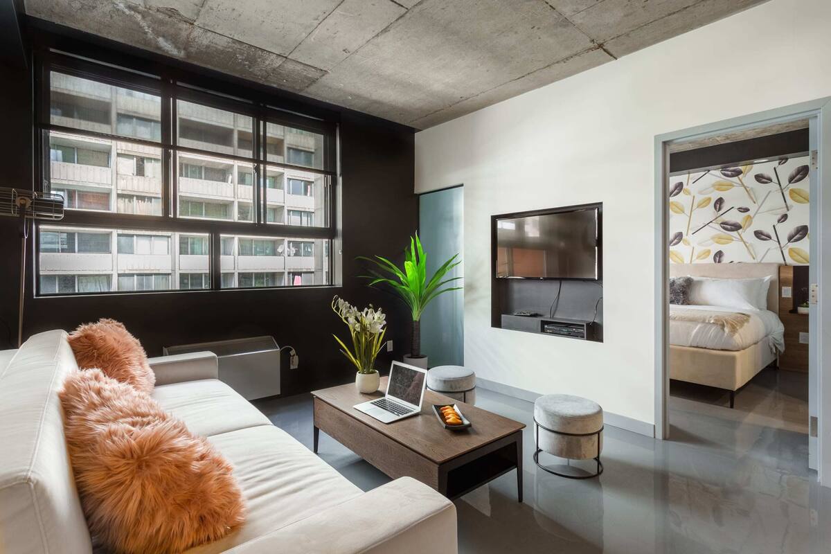 Uma sala de estar moderna com um sofá, mesa de centro e televisão. Há uma janela com vista para edifícios, uma planta e uma mesinha com um laptop e uma bandeja de laranjas.