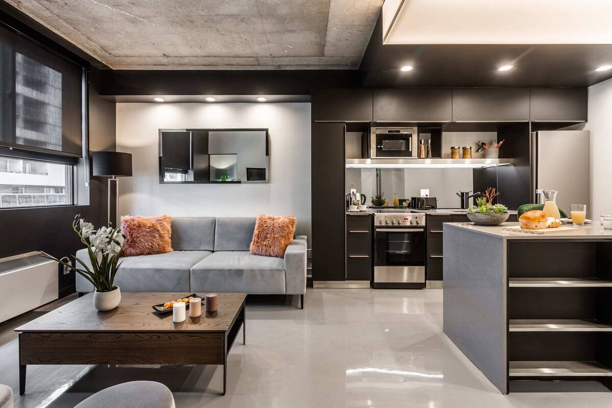 Uma sala de estar moderna com um sofá cinza, uma mesa de centro com objetos decorativos e uma área de cozinha com eletrodomésticos de aço inoxidável.