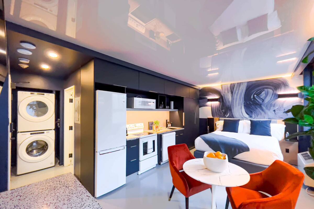 Quarto de hotel moderno com cozinha compacta, cama com roupa de cama azul e branca, uma cadeira vermelha e um quadro na parede.