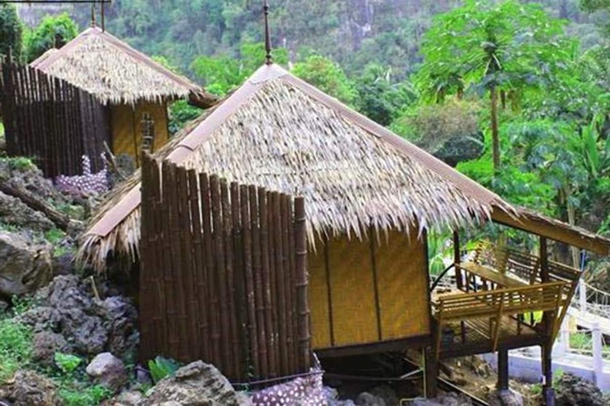 Duas cabanas com telhados de palha e paredes de bambu, localizadas em terreno rochoso com vegetação exuberante ao fundo.
