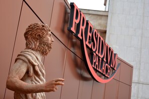 Uma estátua de um homem de barba vestindo uma toga, ao lado de uma placa com a inscrição 'Palácio da Presidência'.
