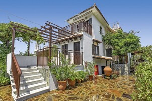 Uma casa de dois andares com um pátio pavimentado em pedra, uma pérgula de madeira e uma escadaria que leva à entrada.
