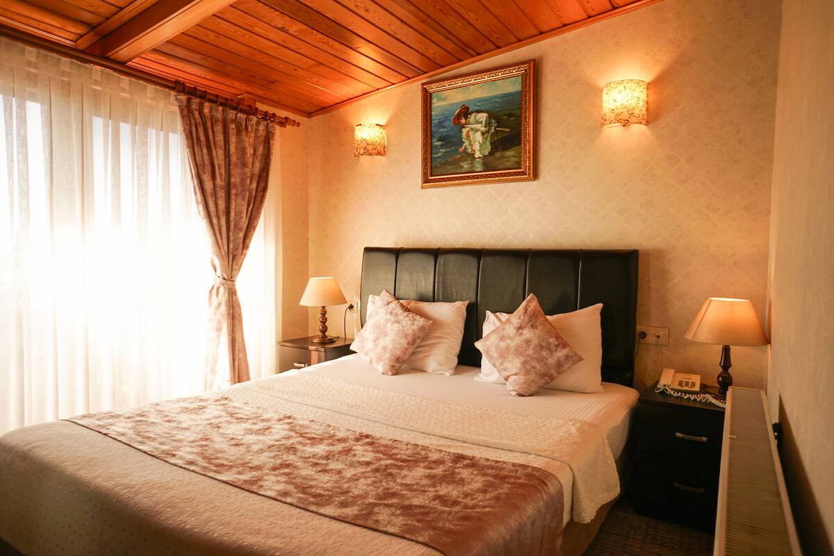 Quarto de hotel com uma cama grande, duas mesas de cabeceira, um criado-mudo, um quadro na parede e uma janela com cortinas.