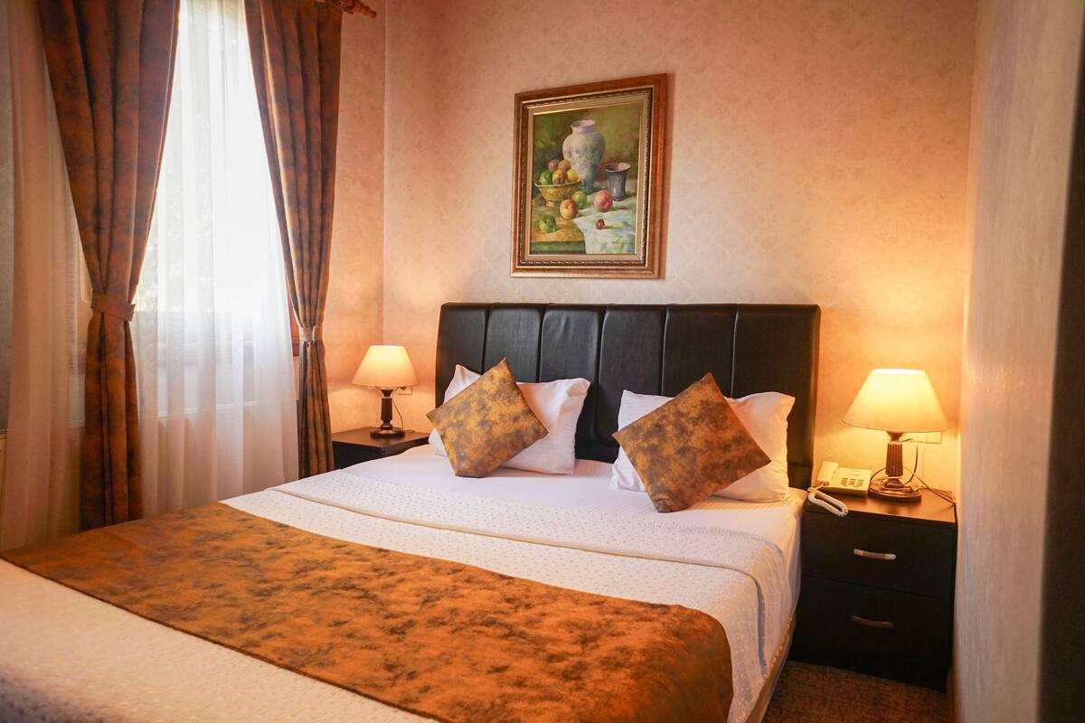 Quarto de hotel com uma cama grande, duas mesas de cabeceira com luminárias, um quadro na parede e uma janela com cortinas.