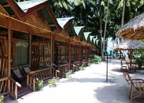Uma fileira de bungalows de madeira com telhados de palha, dispostos em linha ao longo de um caminho de areia, rodeados por palmeiras.