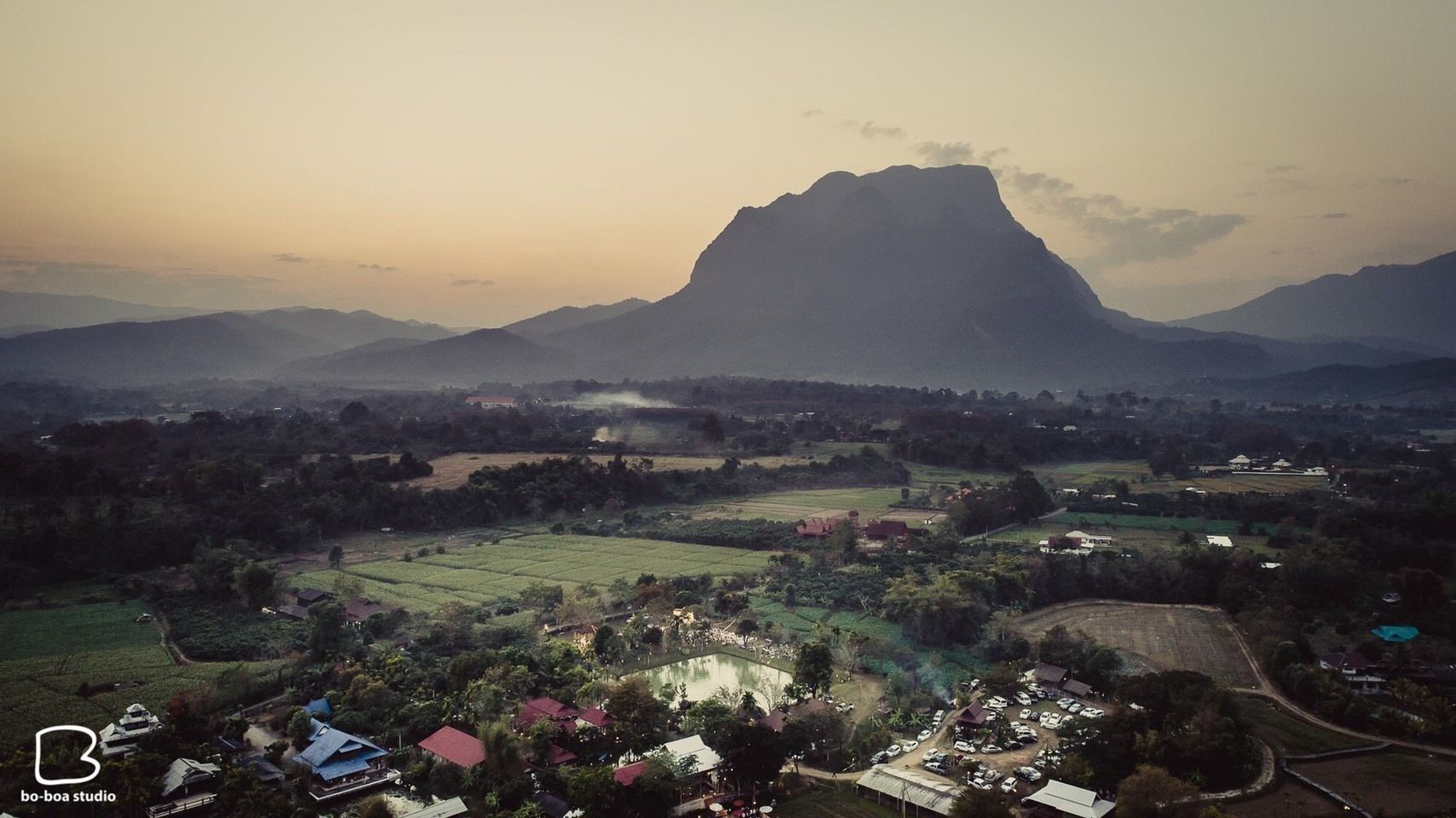Paisagem rural com uma montanha imponente ao fundo, campos e edifícios dispersos.