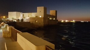 Um edifício costeiro com uma torre imponente, um calçadão à beira-mar e um pôr do sol ao fundo.