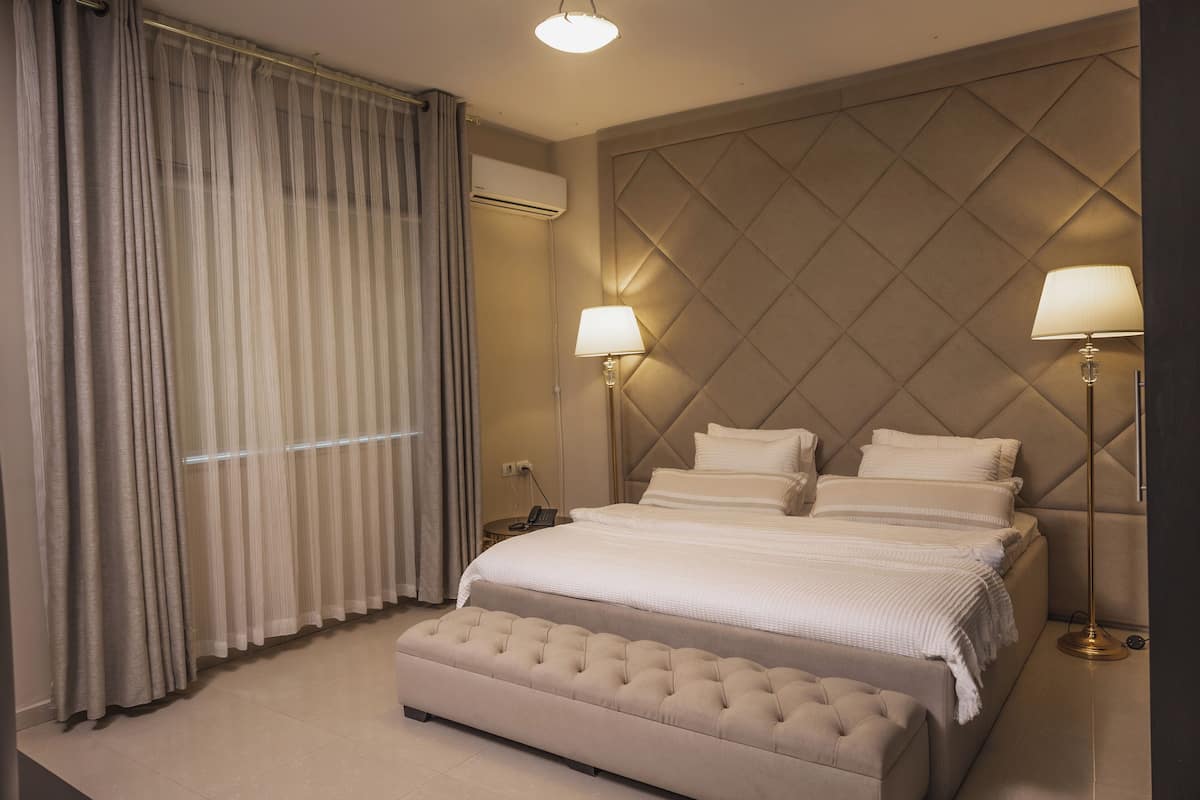 Quarto de hotel com cama grande, duas luminárias de cabeceira, um banco estofado e uma janela com cortinas.