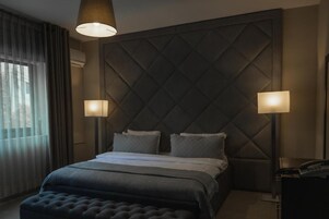Quarto de hotel com uma cama grande, duas luminárias de cabeceira, um painel de cabeceira estofado e uma janela com cortinas.