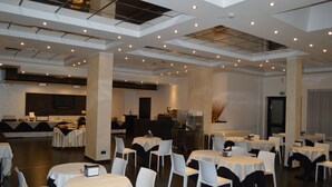 Uma ampla sala de jantar com várias mesas e cadeiras redondas, cozinha ao fundo e design moderno no teto.