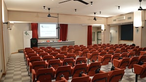 Uma sala de conferências com fileiras de cadeiras laranja, uma tela de projeção e um pódio.