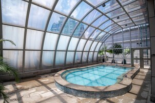 Uma grande piscina interna com teto de vidro, rodeada por piso de pedra e plantas em vasos.