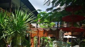 Uma trilha no jardim leva a uma pérgula de madeira com uma placa de boas-vindas, cercada por diversas plantas tropicais e uma fonte de pedra.