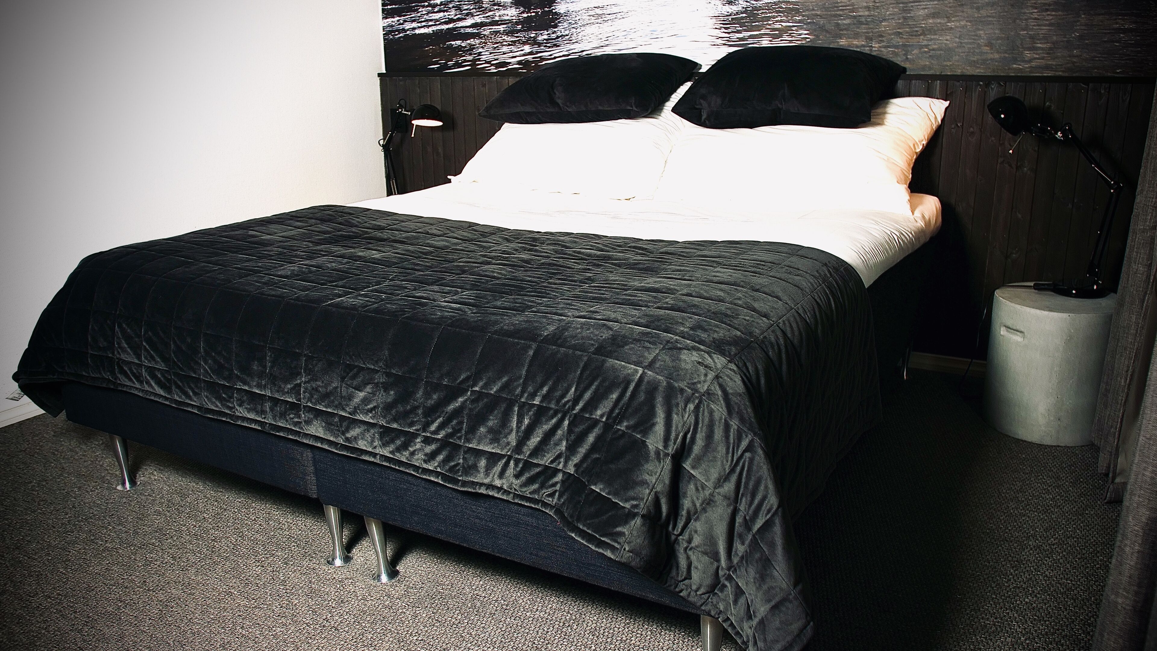 Cama com colcha acolchoada escura, duas almofadas e cabeceira com luminária.