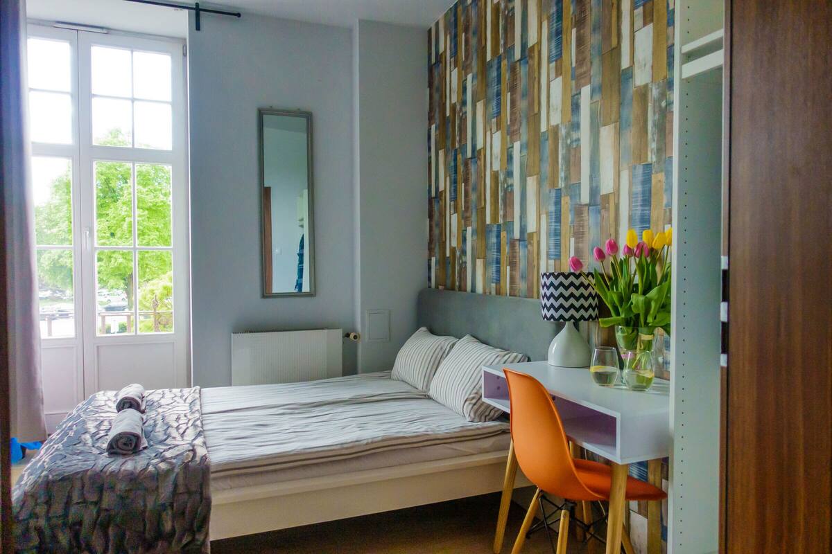 Quarto com uma cama, uma escrivaninha e uma cadeira. Há uma janela com vista para árvores e um carro estacionado lá fora.