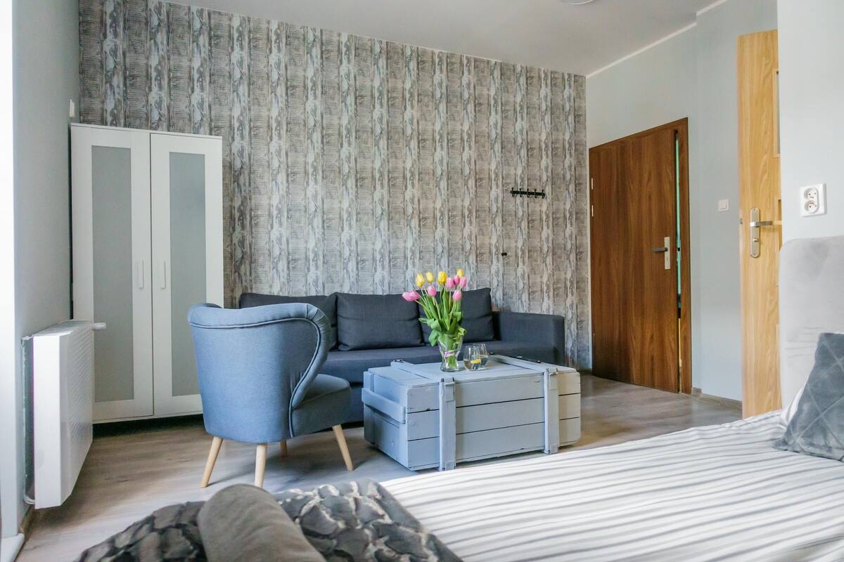 Uma sala de estar moderna com um sofá cinzento, uma poltrona azul, uma mesa de centro e uma cama com roupa de cama listrada.