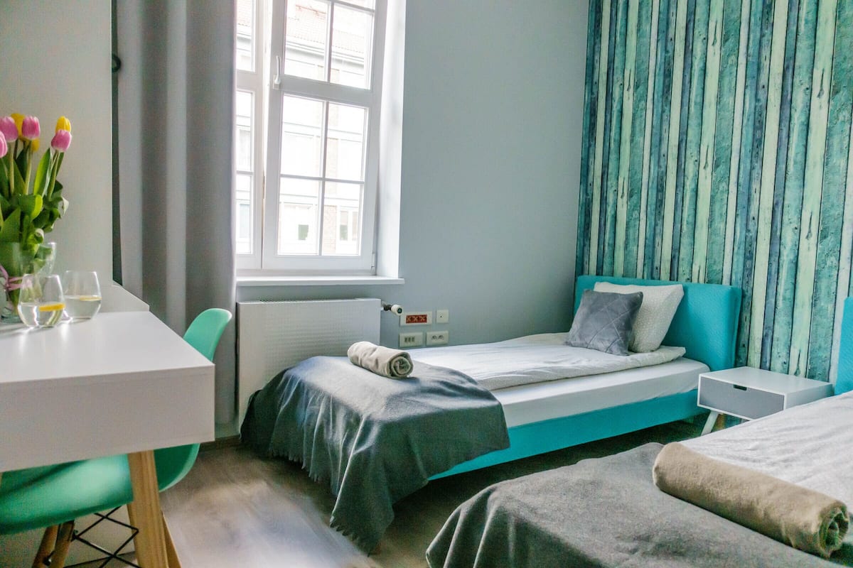 Quarto com cama, mesa de cabeceira, cadeira, janela e parede com linhas verticais.