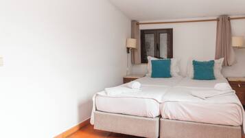 Uma cama de casal com duas almofadas, uma mesa de cabeceira, um abajur e uma janela com cortinas.