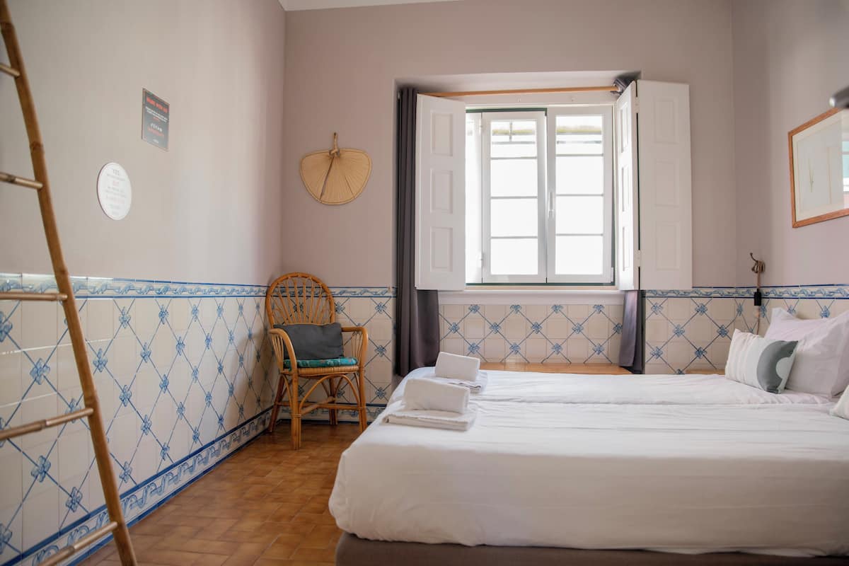 Quarto de hotel com duas camas, uma cadeira, uma escada e uma janela com cortinas.