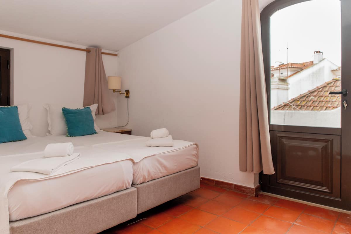 Quarto de hotel com cama, mesa de cabeceira, abajur e vista para edifícios através de uma porta.
