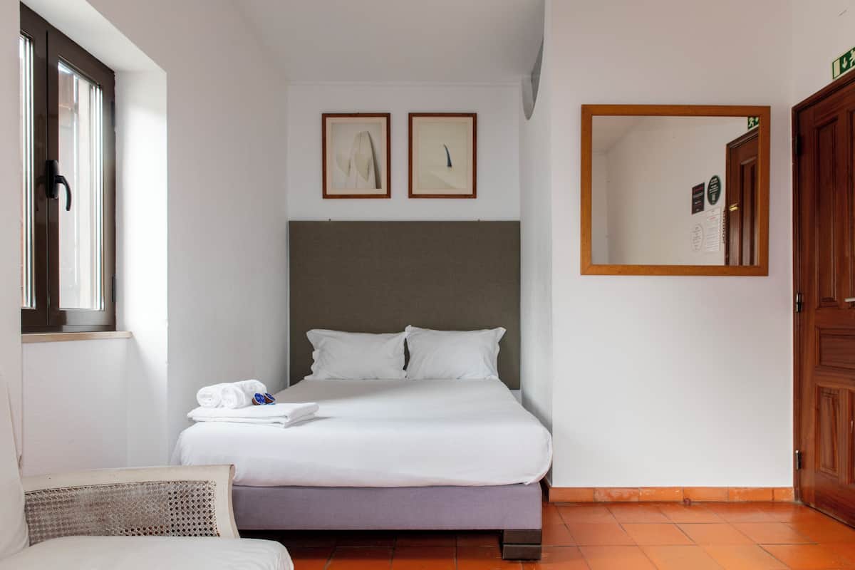 Quarto de hotel com uma cama, uma cadeira, uma janela e duas imagens emolduradas na parede.