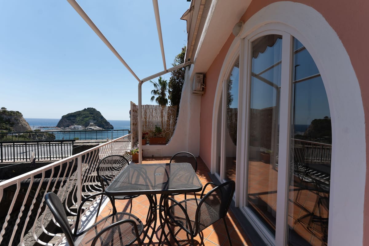 Uma varanda com mesa e cadeiras, guarda-corpo e vista para o mar e uma ilha rochosa.