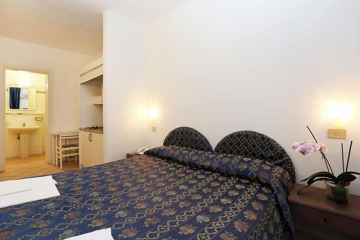 Quarto com cama, mesa de cabeceira e uma pequena escrivaninha com cadeira. Banheiro visível por uma porta aberta.