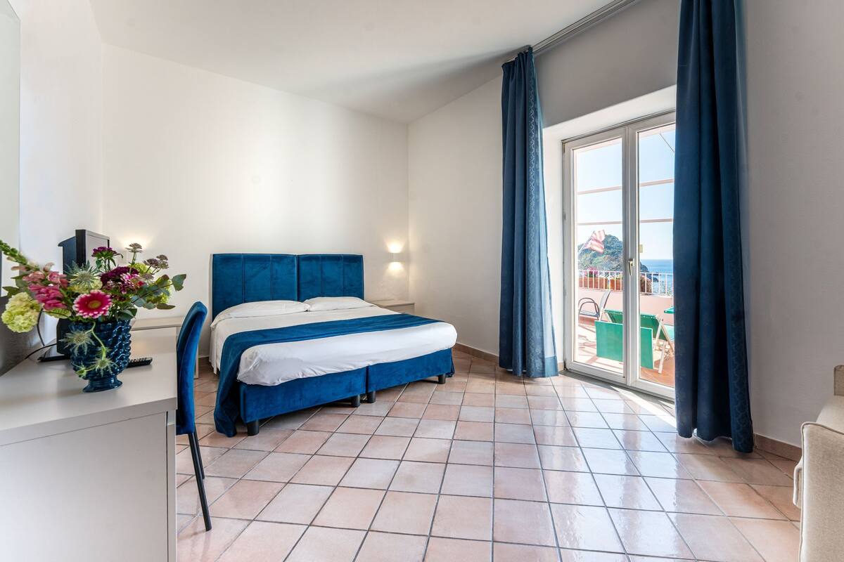 Quarto de hotel com uma cama, uma escrivaninha com um vaso de flores, uma cadeira e vista pela janela grande.