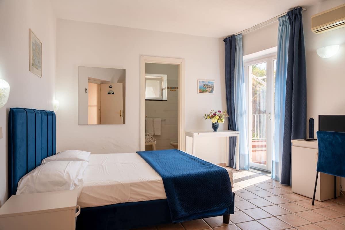 Quarto com cama, escrivaninha, cadeira, janela com cortinas, porta que dá para um banheiro e uma pequena mesa com um vaso de flores.