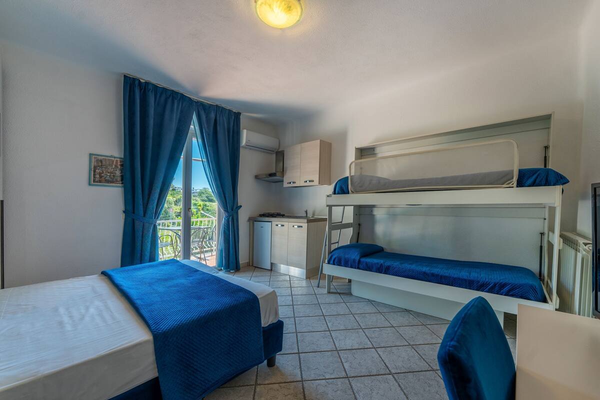 Quarto com duas camas, uma cozinha compacta e uma janela com cortinas.