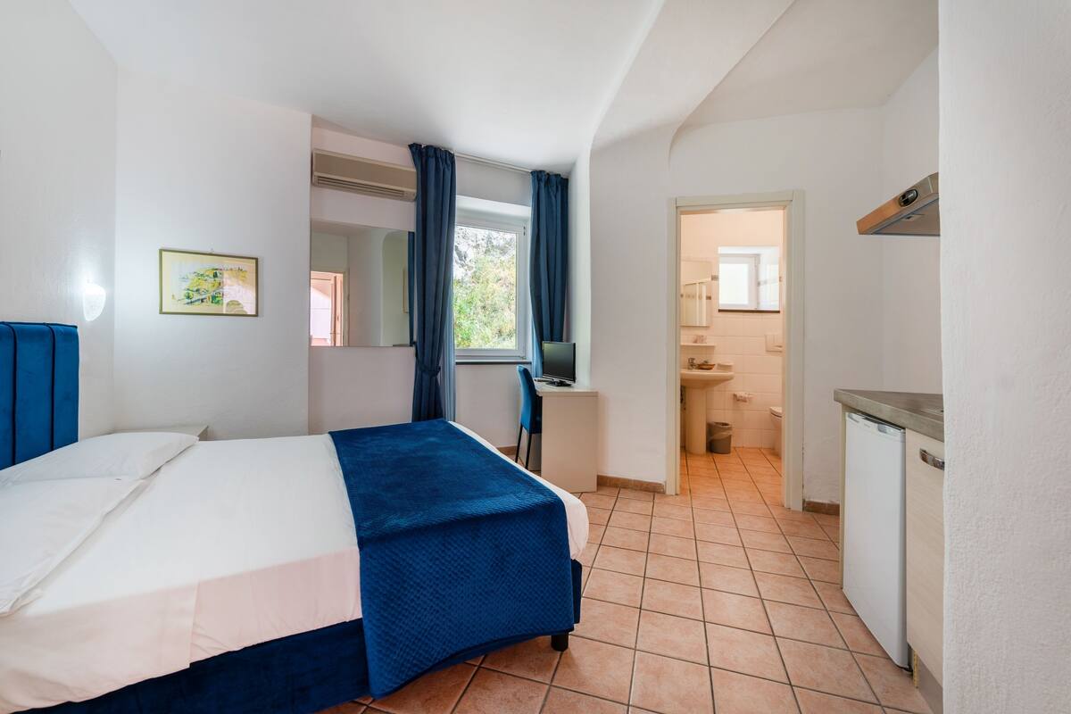 Quarto com cama, escrivaninha, televisão e banheiro visível por uma porta aberta.