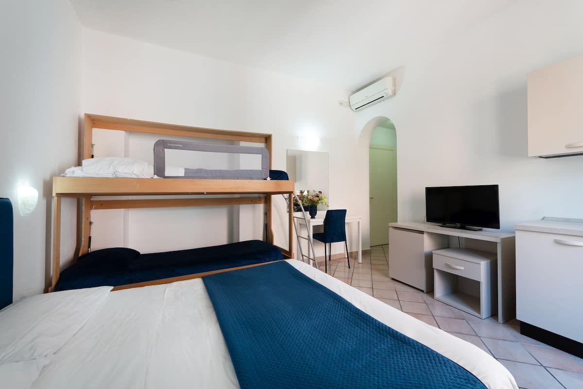 Quarto de hotel com uma cama de solteiro e beliche, uma mesa com televisão, uma cadeira e um pequeno armário.