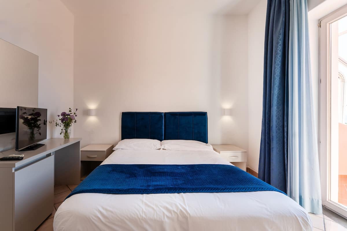 Quarto de hotel moderno com cama, mesas de cabeceira, televisão e janela com cortinas.