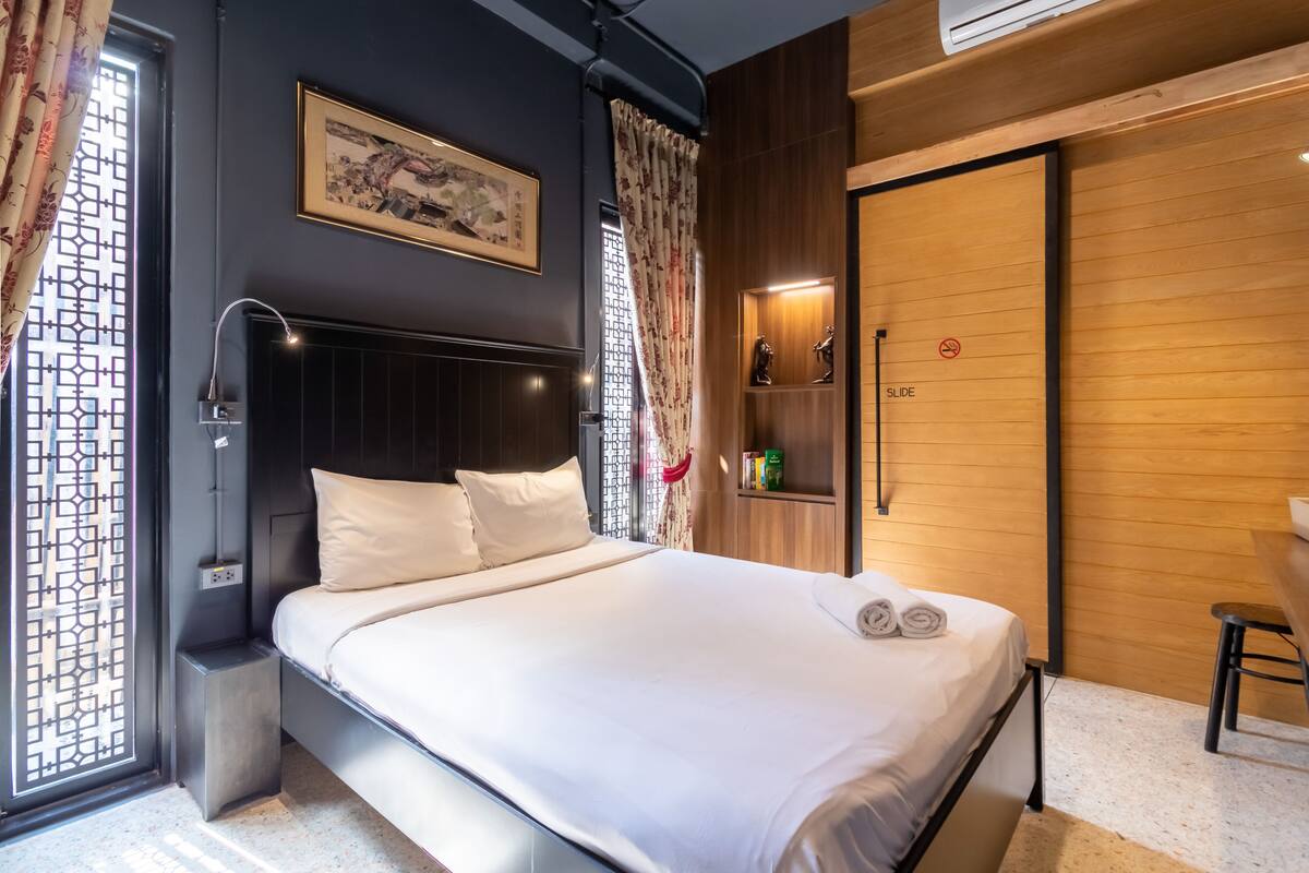 Quarto de hotel moderno com uma cama grande, mesa de cabeceira, armário de madeira e um pequeno espaço para sentar.
