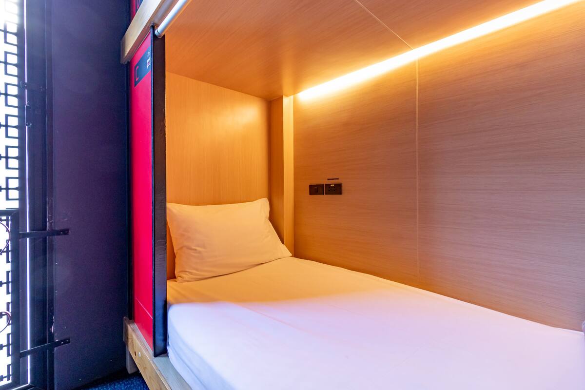 Quarto de hotel compacto com uma cama de solteiro, cabeceira de madeira e uma parede com detalhes em vermelho.