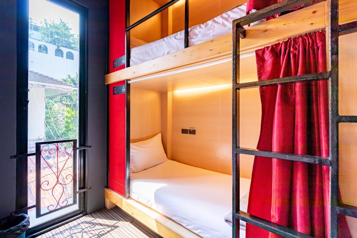 Quarto com beliche, janela, cortina vermelha e vista para um edifício.