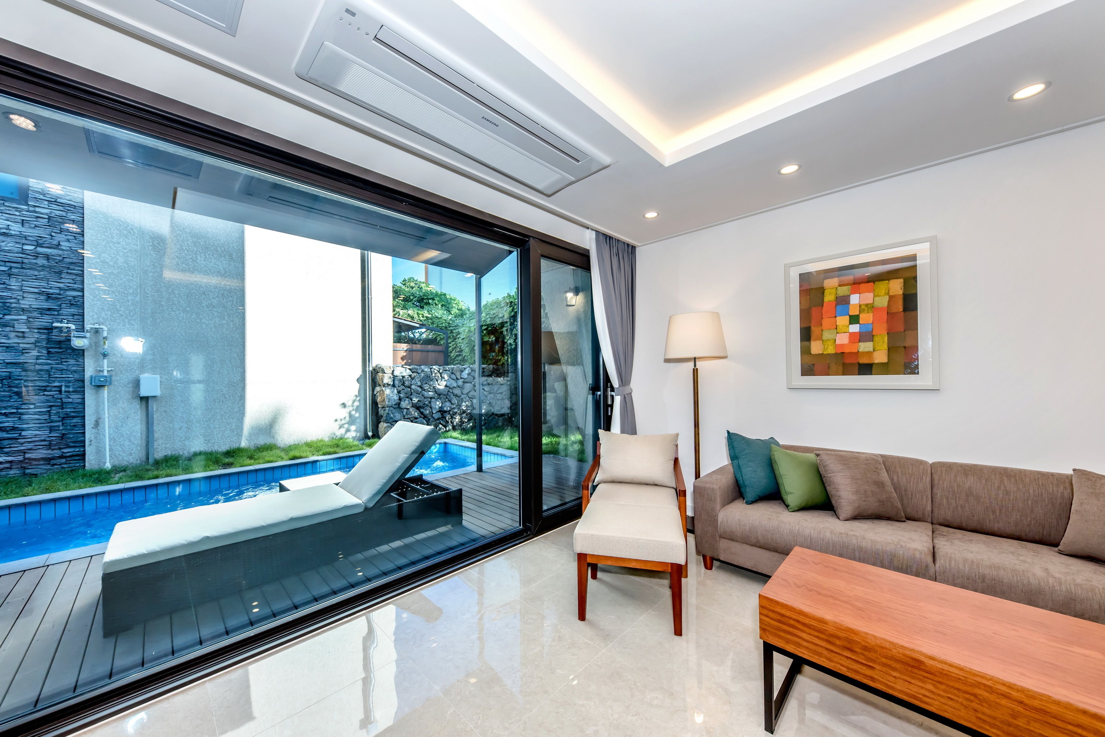Uma sala de estar moderna com parede de vidro, piscina, sofá e mesa de centro de madeira.