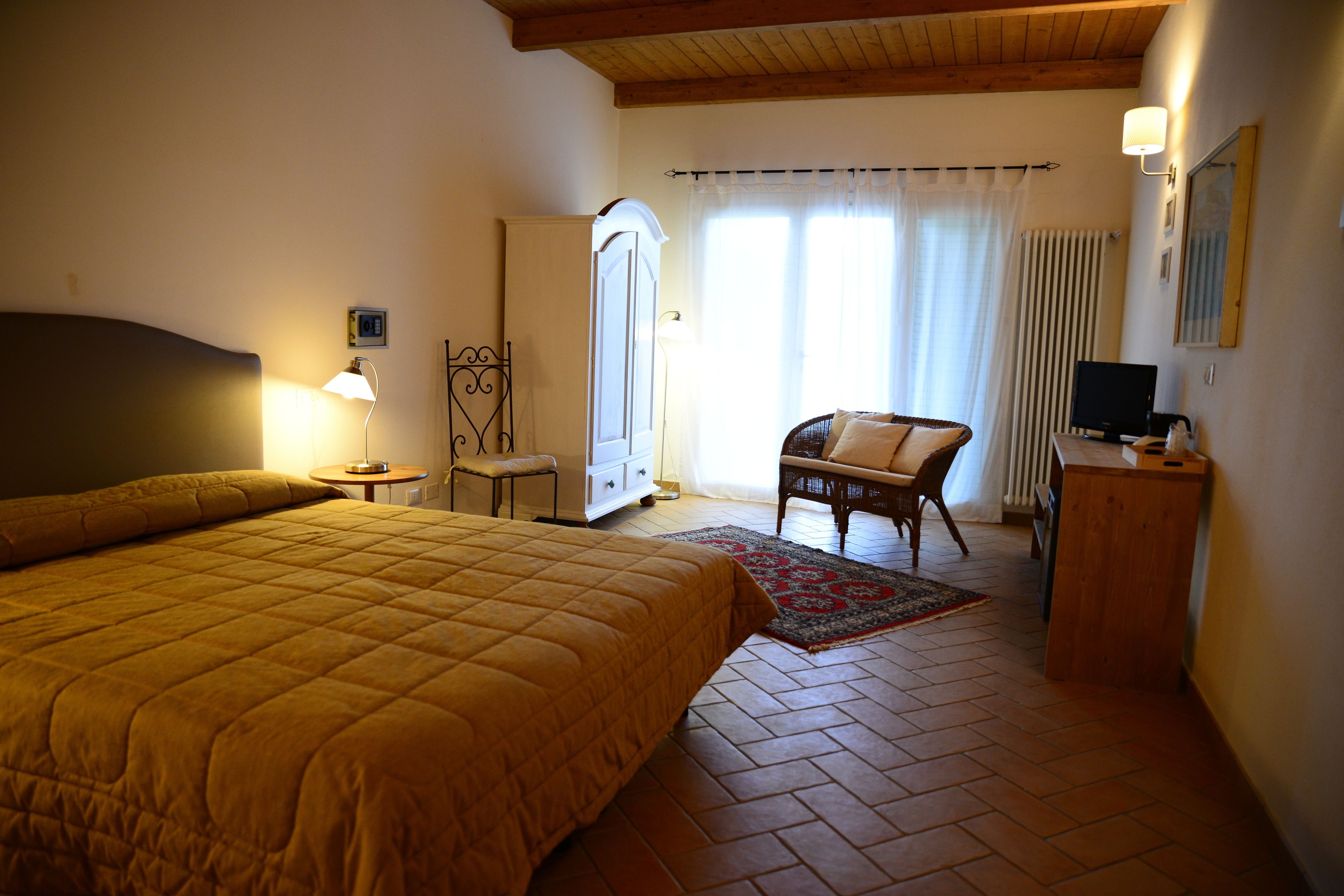 Quarto de hotel com uma cama grande, uma cadeira, uma escrivaninha com computador e uma janela com cortinas.