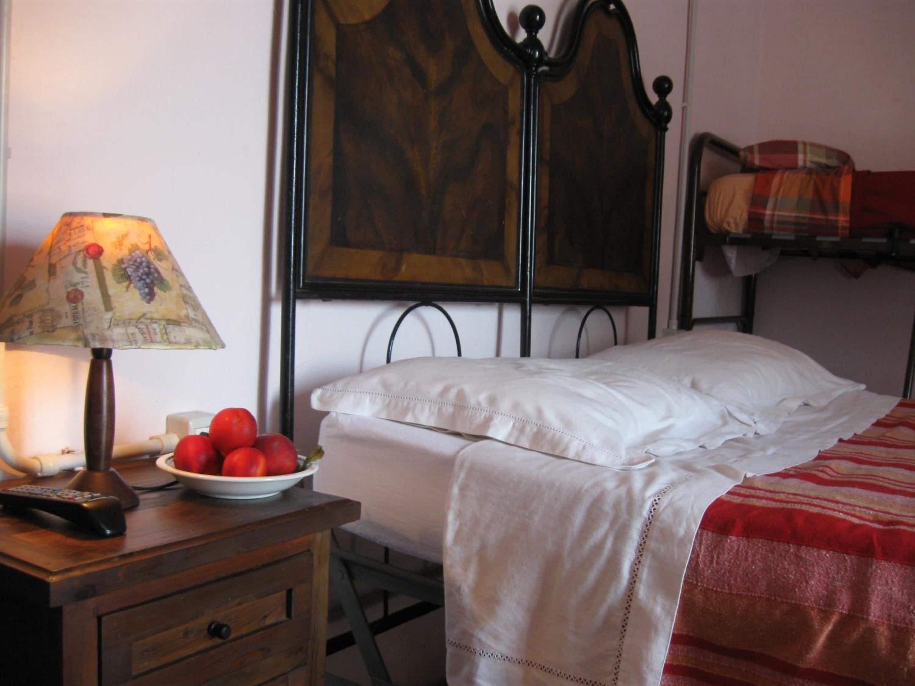 Quarto com cama, mesa de cabeceira com abajur, uma tigela de frutas e um quadro na parede.