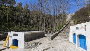 Uma escadaria de pedra sobe uma colina, ladeada por paredes brancas com portas azuis, e uma pequena construção com uma porta amarela na base.