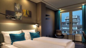 Quarto de hotel com uma cama grande, uma cadeira, uma mesinha e vista para edifícios pela janela.