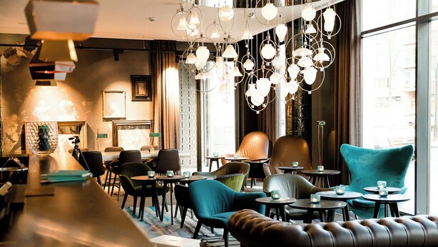 Interior moderno com área de bar, assentos com mesas redondas e uma grande janela que traz luz natural.