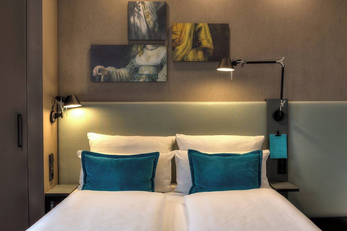 Quarto de hotel com uma cama, duas mesas de cabeceira, um abajur fixado na parede e três quadros.