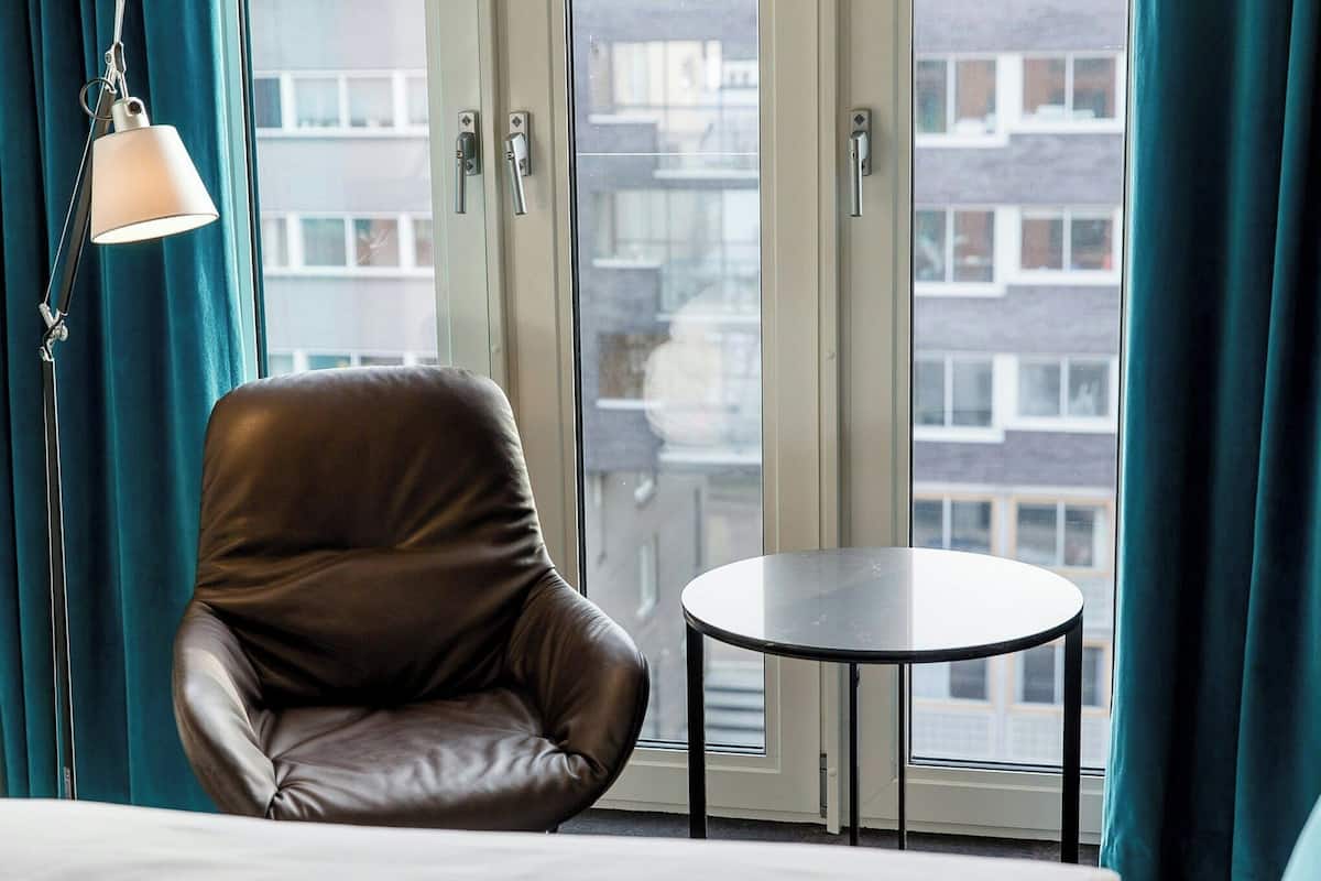 Quarto com poltrona de couro, mesa redonda pequena e janela com cortinas.