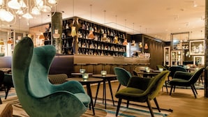 Uma área de bar moderna com cadeiras estofadas em tons de verde-água e verde, diversas garrafas em prateleiras e iluminação ambiente.