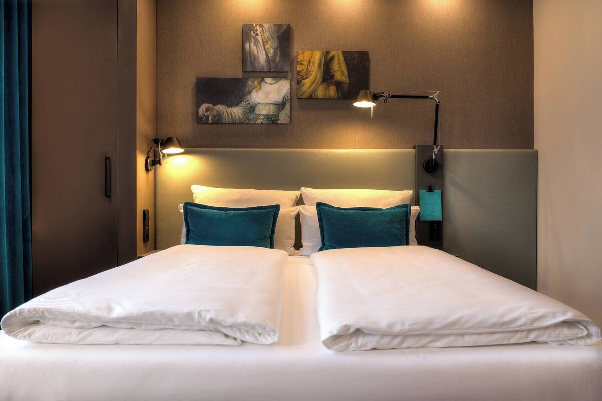 Uma cama de casal com roupa de cama branca, almofadas verde-água e duas luminárias de parede para leitura.