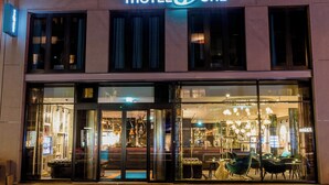 Um motel moderno com um letreiro luminoso, amplas janelas de vidro e um interior visível, com móveis e iluminação.